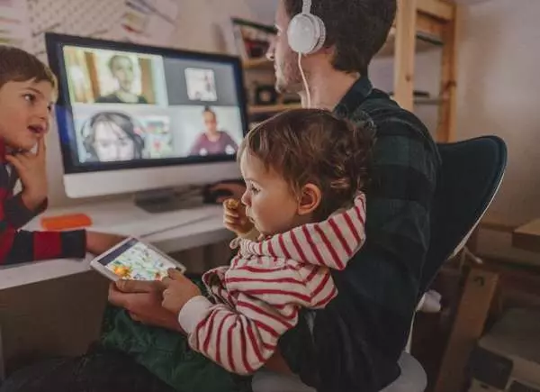 Pria di komputer, melihat layar sambil menggendong satu bayi di pangkuannya dan membantu balita lain dengan tablet