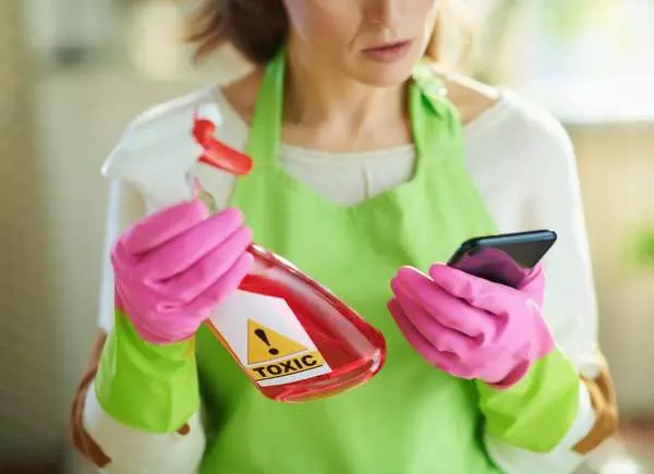 Vrouw in schort met fles reiniger met gif erop geschreven; ze kijkt naar iets op haar telefoon.
