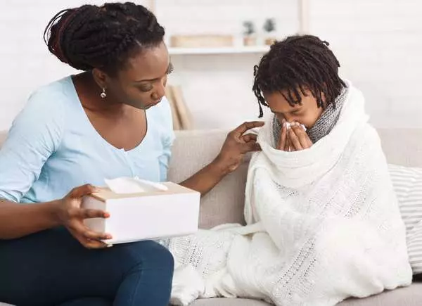 Černá žena sedí s černou dívkou na gauči; dívka je zabalená do deky a kýchá