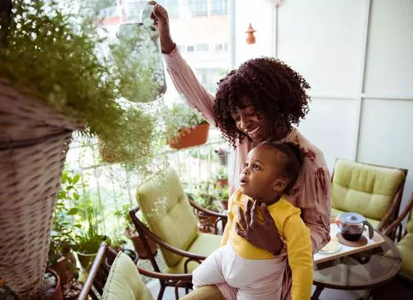 Czarna kobieta trzyma dziecko podczas podlewania roślin w pomieszczeniu.