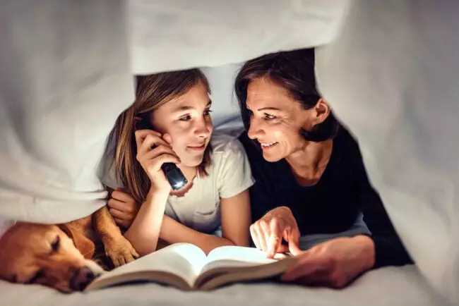 Mutter, Tochter und Hund, die spät in der Nacht unter der Decke auf dem Bett liegen und eine Taschenlampe halten und ein Buch lesen